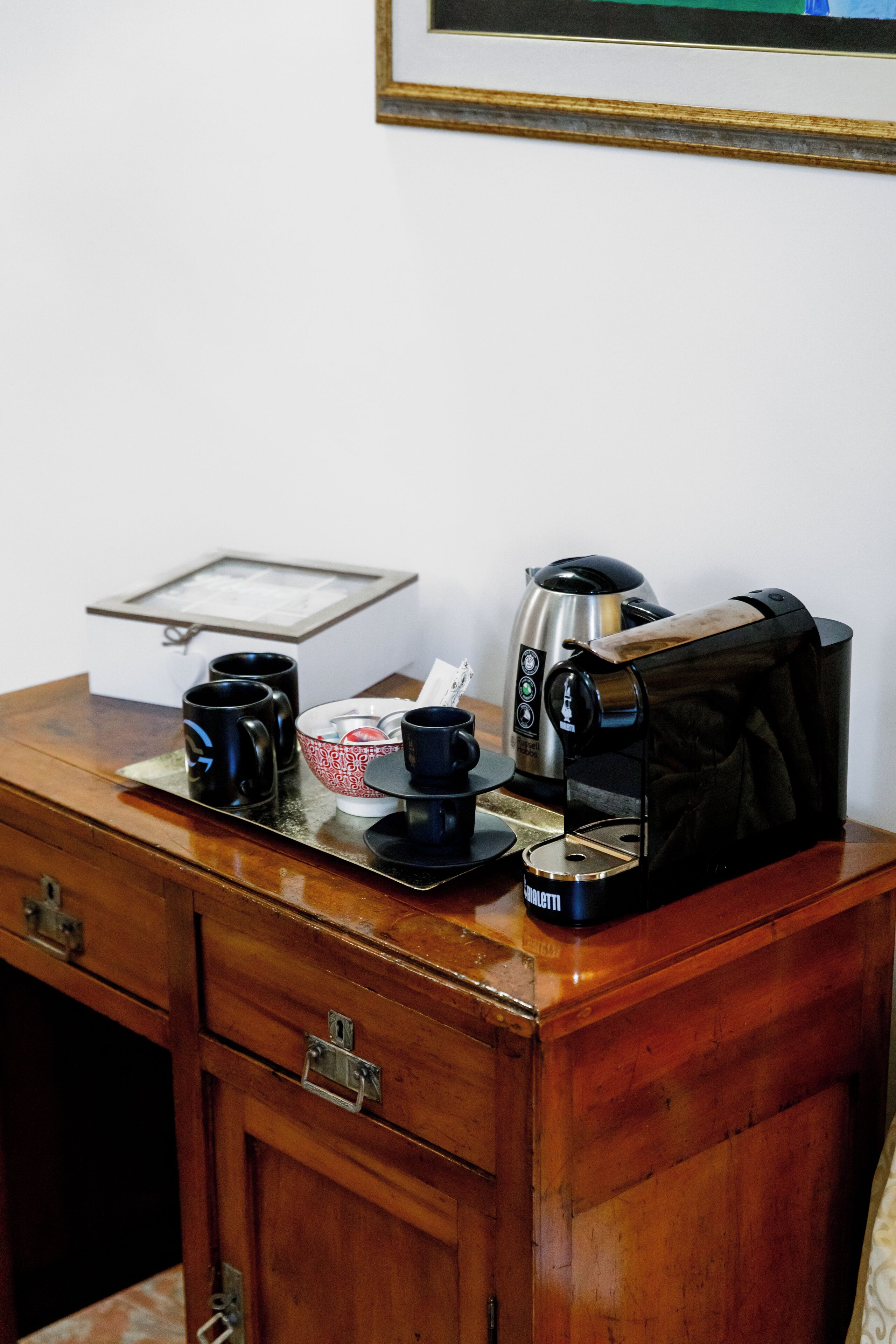 Uma secretária de madeira com uma máquina de café, um quadro emoldurado e uma bandeja com chávenas e uma caixa.