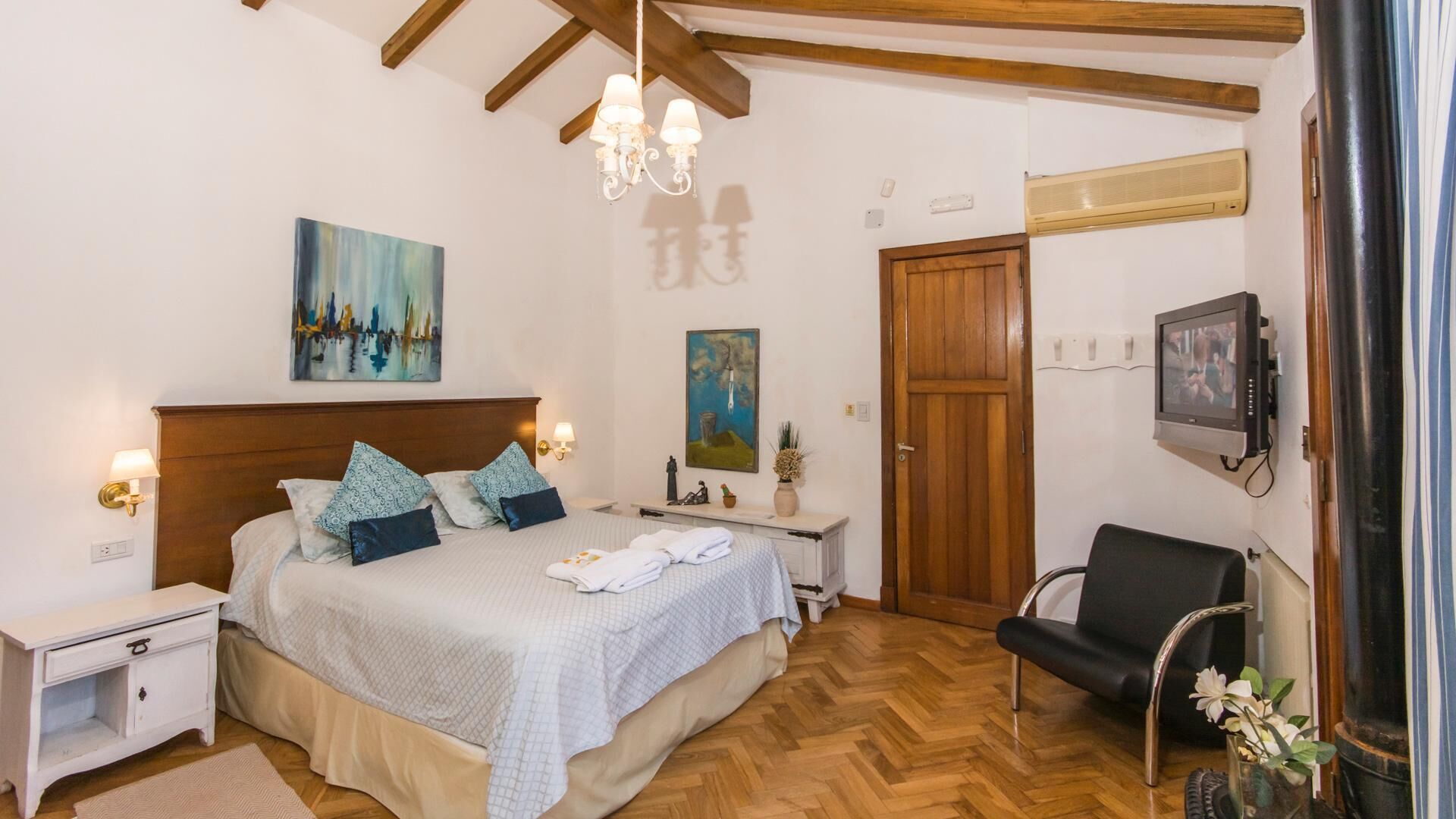 Quarto com cama de madeira, mesas de cabeceira, uma cadeira, televisão e obras de arte nas paredes.