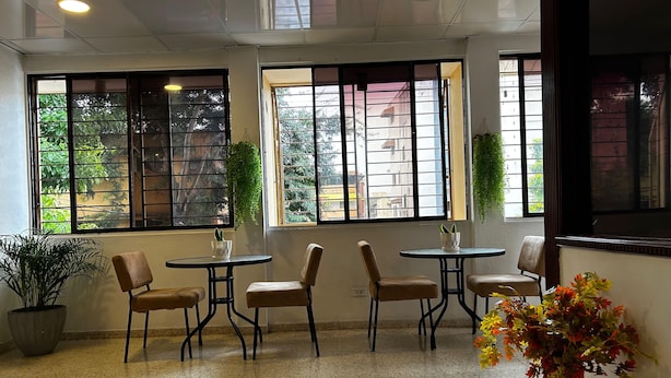 Um quarto com quatro mesas e cadeiras, janelas amplas com grades de metal e plantas em vasos.