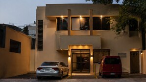 Um edifício moderno com entrada de garagem, dois carros estacionados e uma árvore à direita.