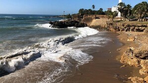 Uma praia com ondas quebrando na costa, um afloramento rochoso e um edifício branco com telhado vermelho ao fundo.