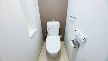 Casa de banho pequena e moderna com sanita branca, suporte de papel higiénico e um botão de descarga na parede.