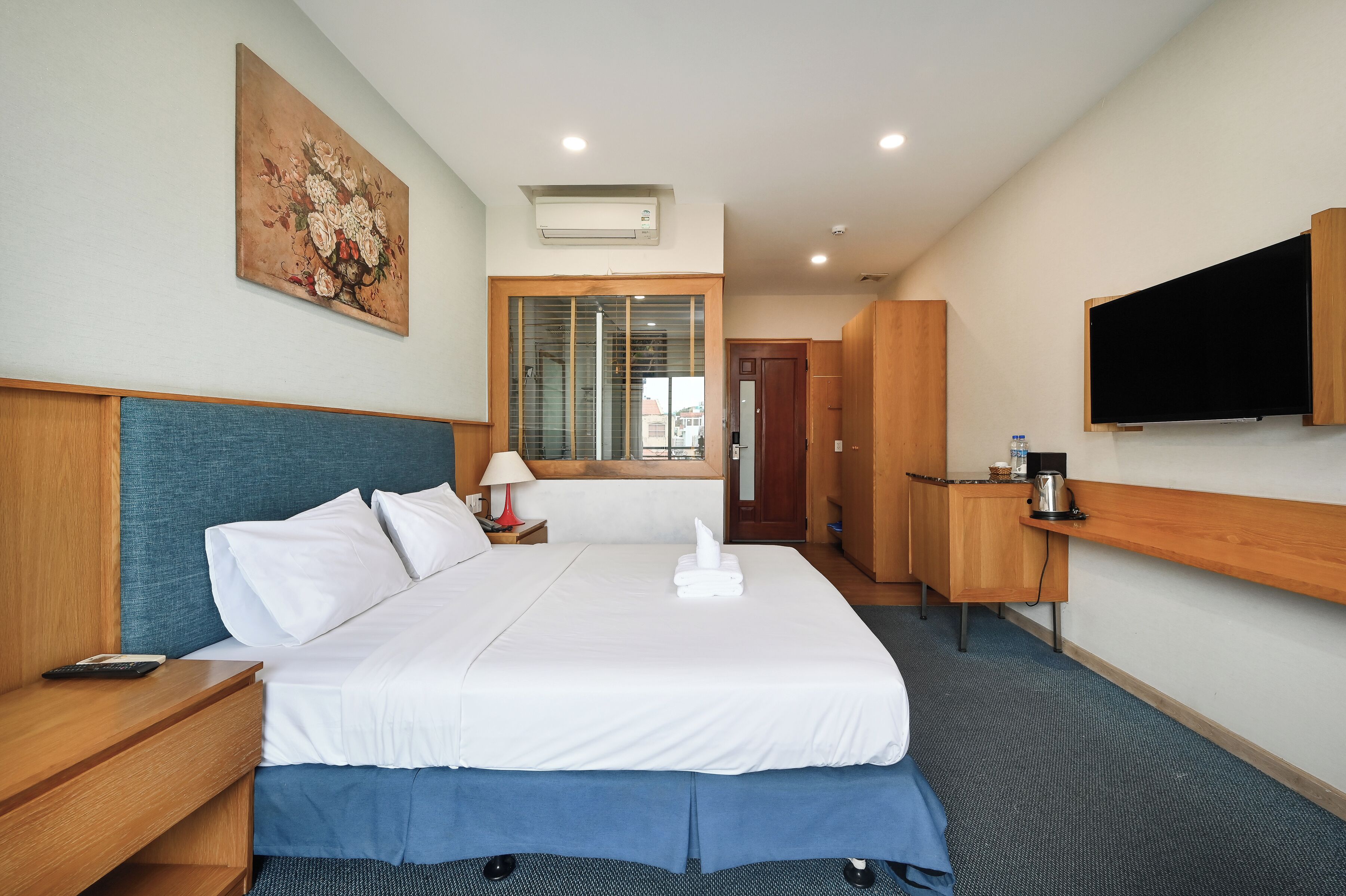 Quarto de hotel com uma cama grande, cabeceira de madeira, TV de tela plana na parede, escrivaninha de madeira com luminária e um quadro na parede.