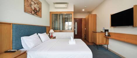 Quarto de hotel com uma cama grande, cabeceira de madeira, TV de tela plana na parede, escrivaninha de madeira com luminária e um quadro na parede.
