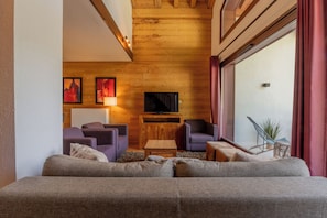 Uma sala de estar aconchegante com teto de madeira, um sofá, duas poltronas, uma mesa de centro, uma TV e uma janela com cortinas.