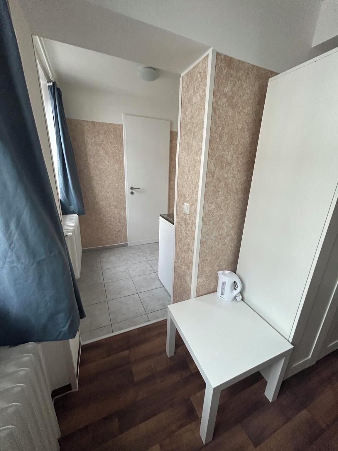 Quarto de hotel com uma cama, uma pequena mesa com uma chávena, um radiador e uma porta que dá para outro quarto.