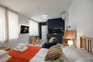Quarto de hotel com cama, escrivaninha, cadeira, cozinha compacta e televisão.