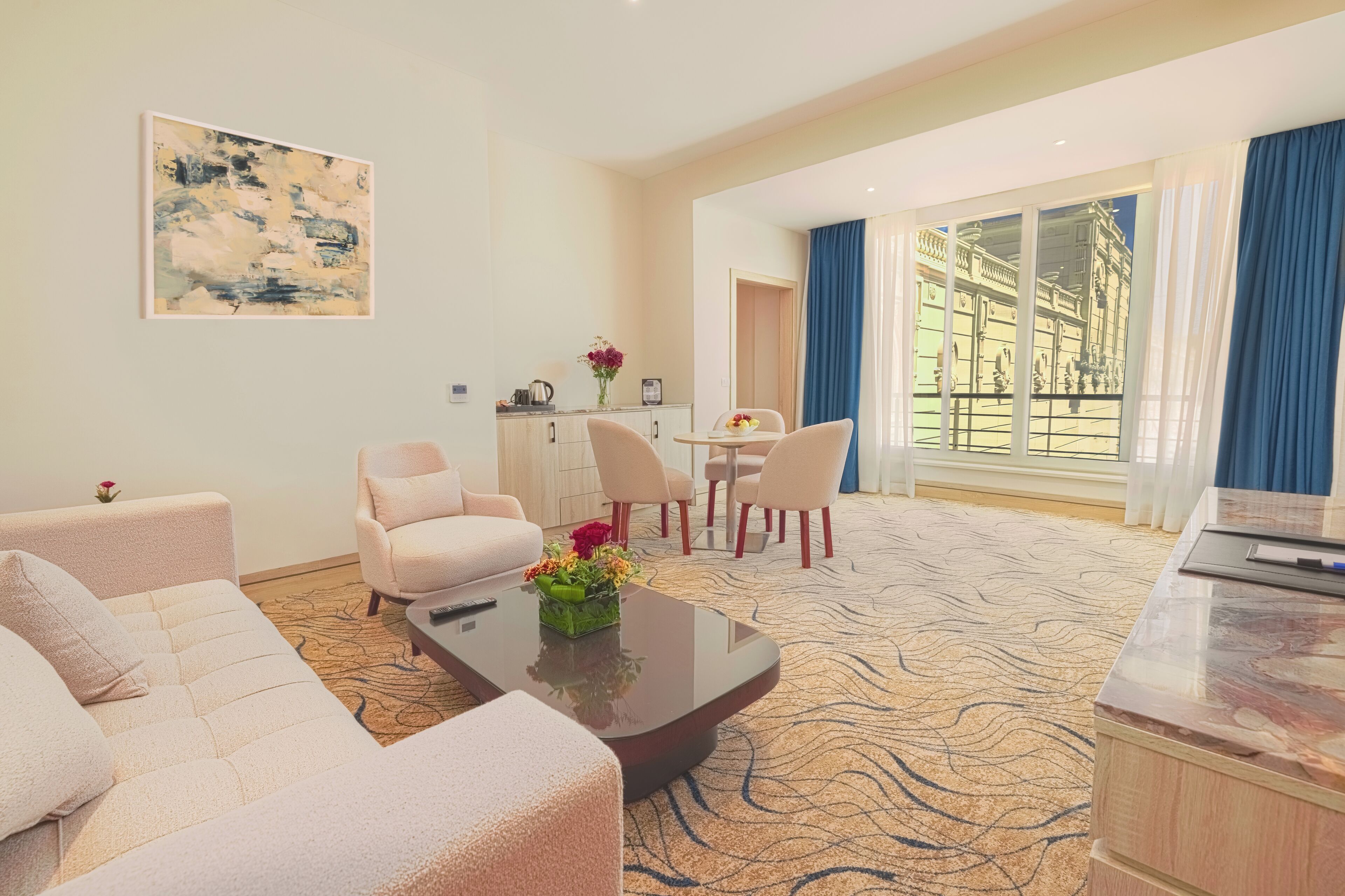 Quarto de hotel moderno com sofá, mesa de centro com flores, área de refeições com cadeiras e vista para um edifício através de grandes janelas.