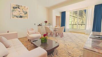 Quarto de hotel moderno com sofá, mesa de centro com flores, área de refeições com cadeiras e vista para um edifício através de grandes janelas.