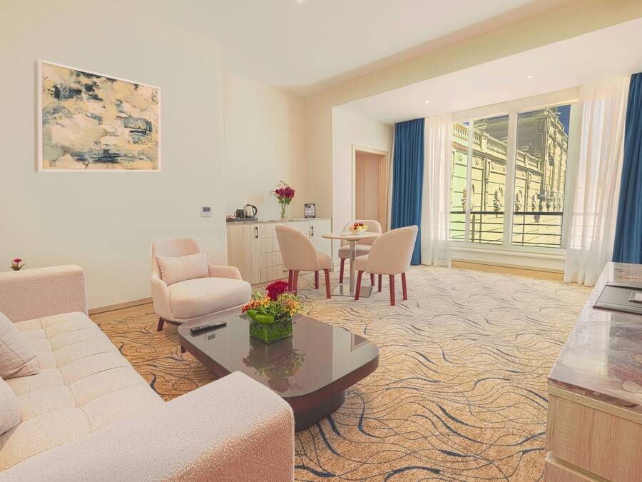 Quarto de hotel moderno com sofá, mesa de centro com flores, área de refeições com cadeiras e vista para um edifício através de grandes janelas.