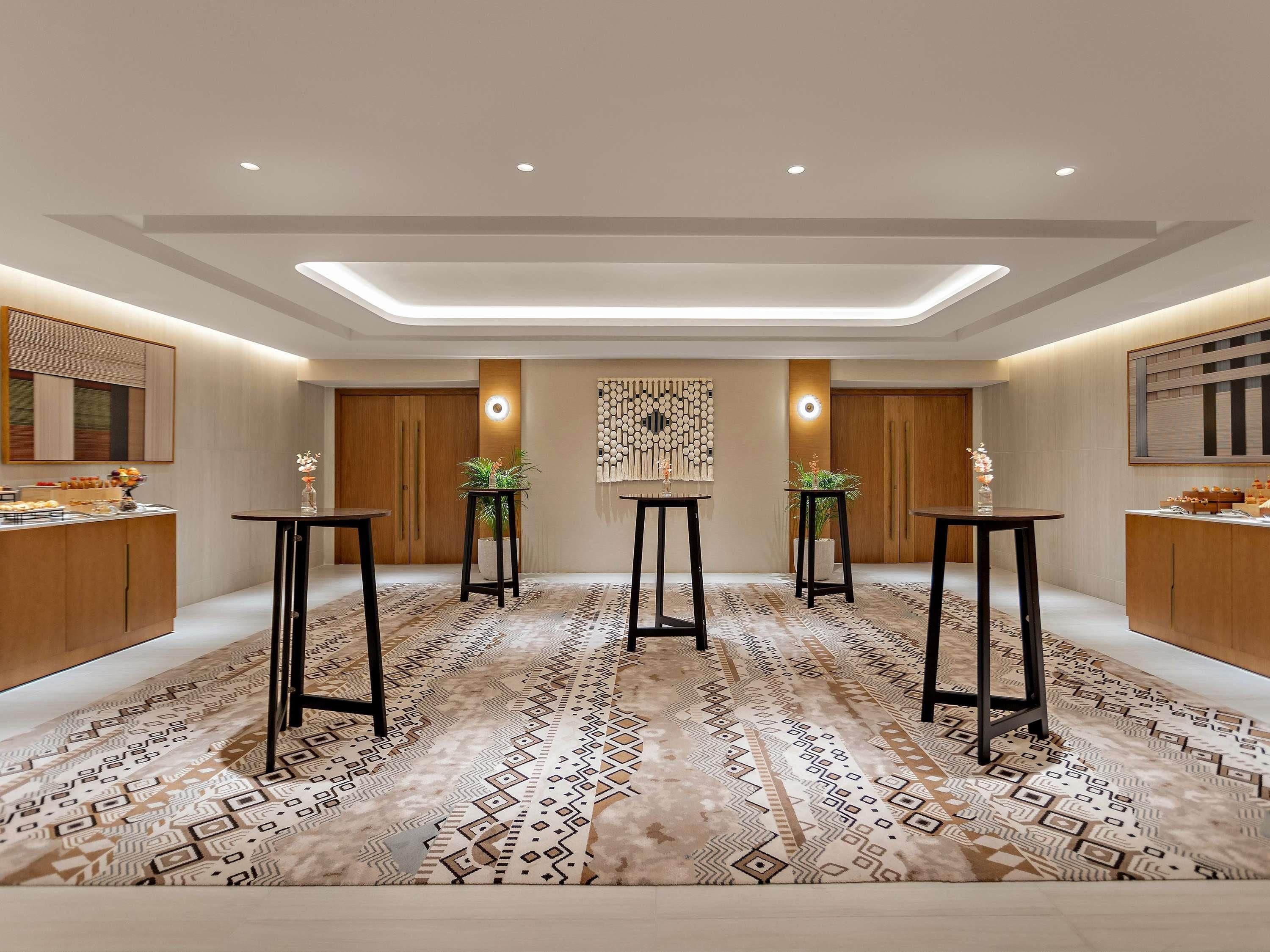 Um lobby de hotel moderno com carpete estampado, armários de madeira e três mesas pretas com plantas em vasos.