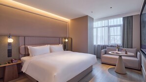 Quarto de hotel moderno com uma cama grande, um sofá, uma mesinha redonda e uma janela com cortinas.
