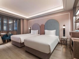 Quarto de hotel com duas camas, área de estar com uma poltrona, uma mesa pequena e uma mesa de cabeceira.
