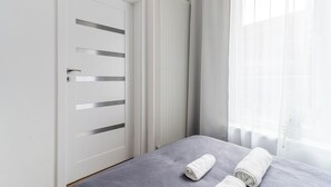 Quarto com cama, porta branca com ripas horizontais, radiador e cortinas leves.