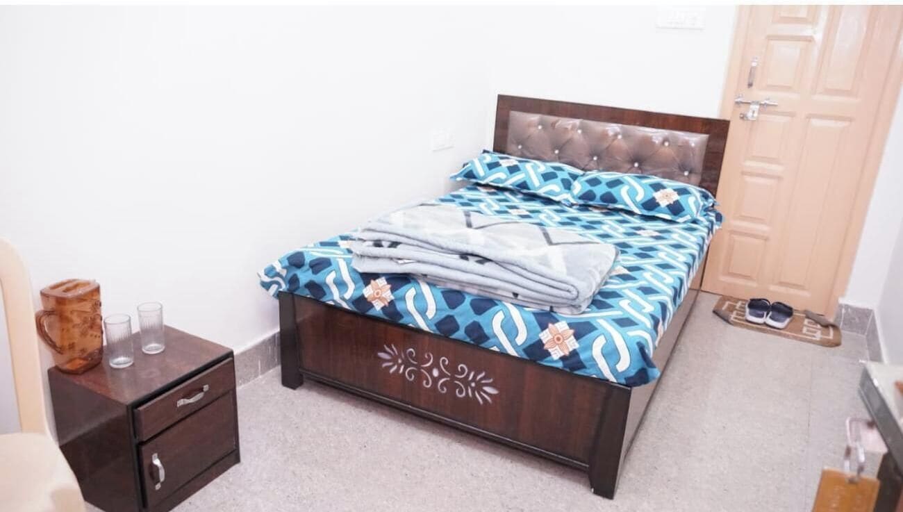 Uma cama bem arrumada com roupa de cama estampada, um criado-mudo de madeira com gaveta e uma porta ao fundo.