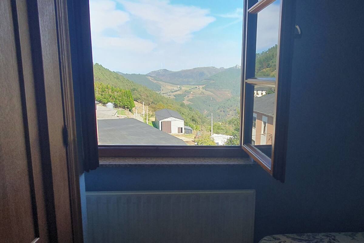 Quarto com janela dando vista para uma paisagem montanhosa com edifícios e turbinas eólicas.
