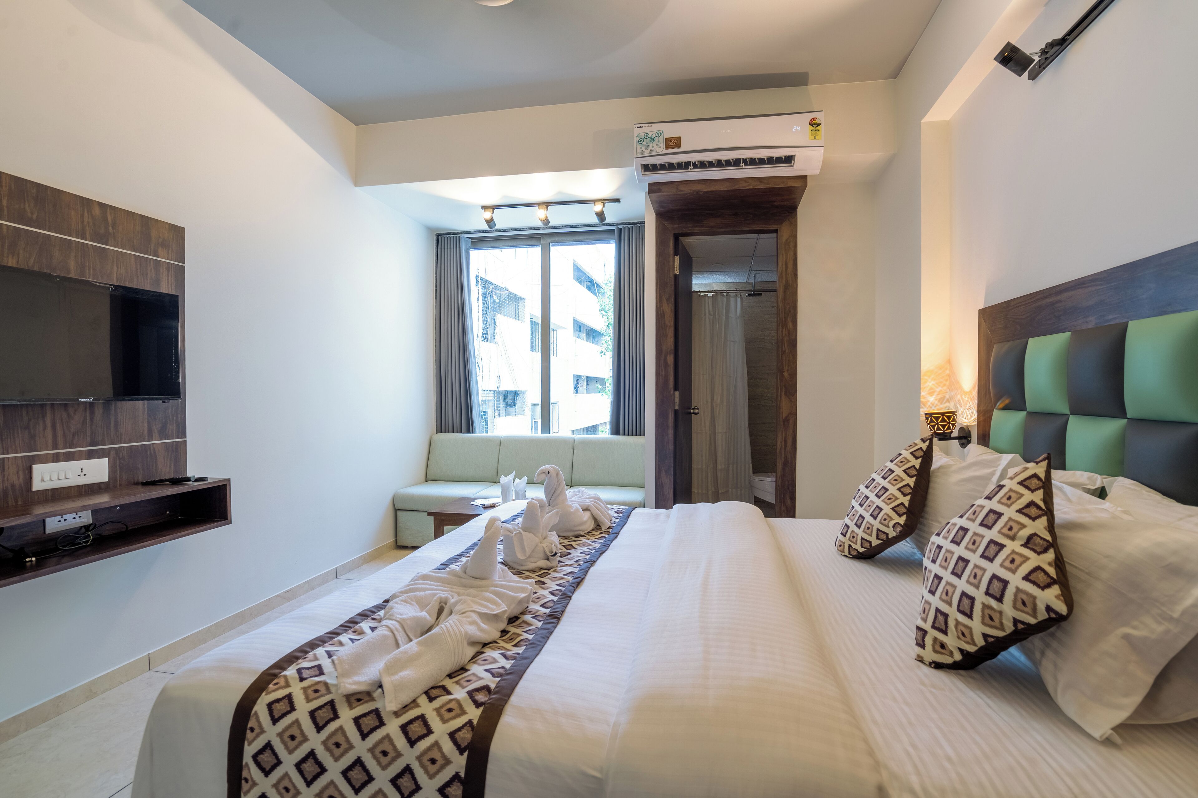 Quarto de hotel com cama, televisão fixada na parede, janela com cortinas e unidade de ar condicionado.