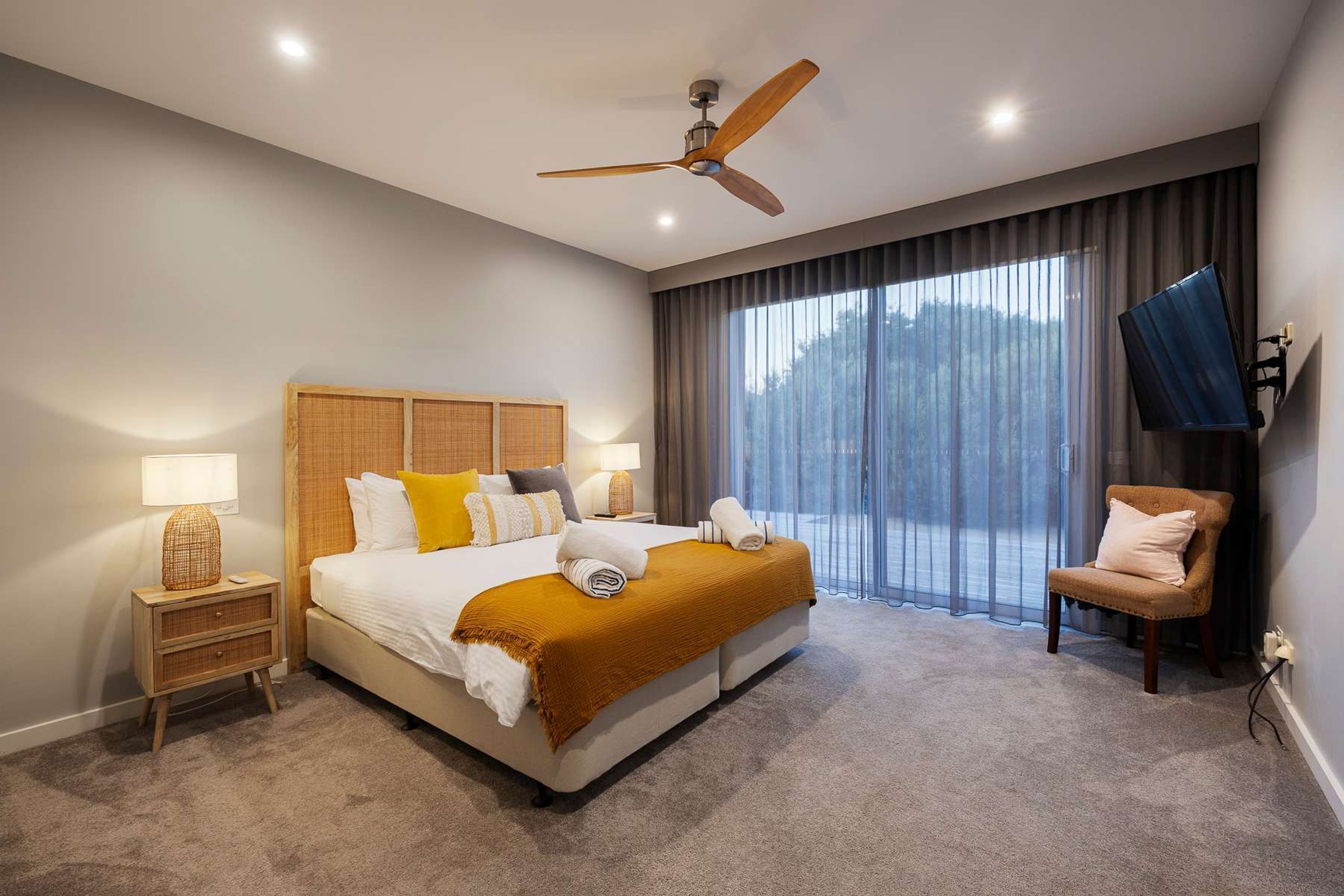 Quarto de hotel moderno com uma cama grande, ventilador de teto, TV fixada na parede e vista para o exterior através de amplas janelas.