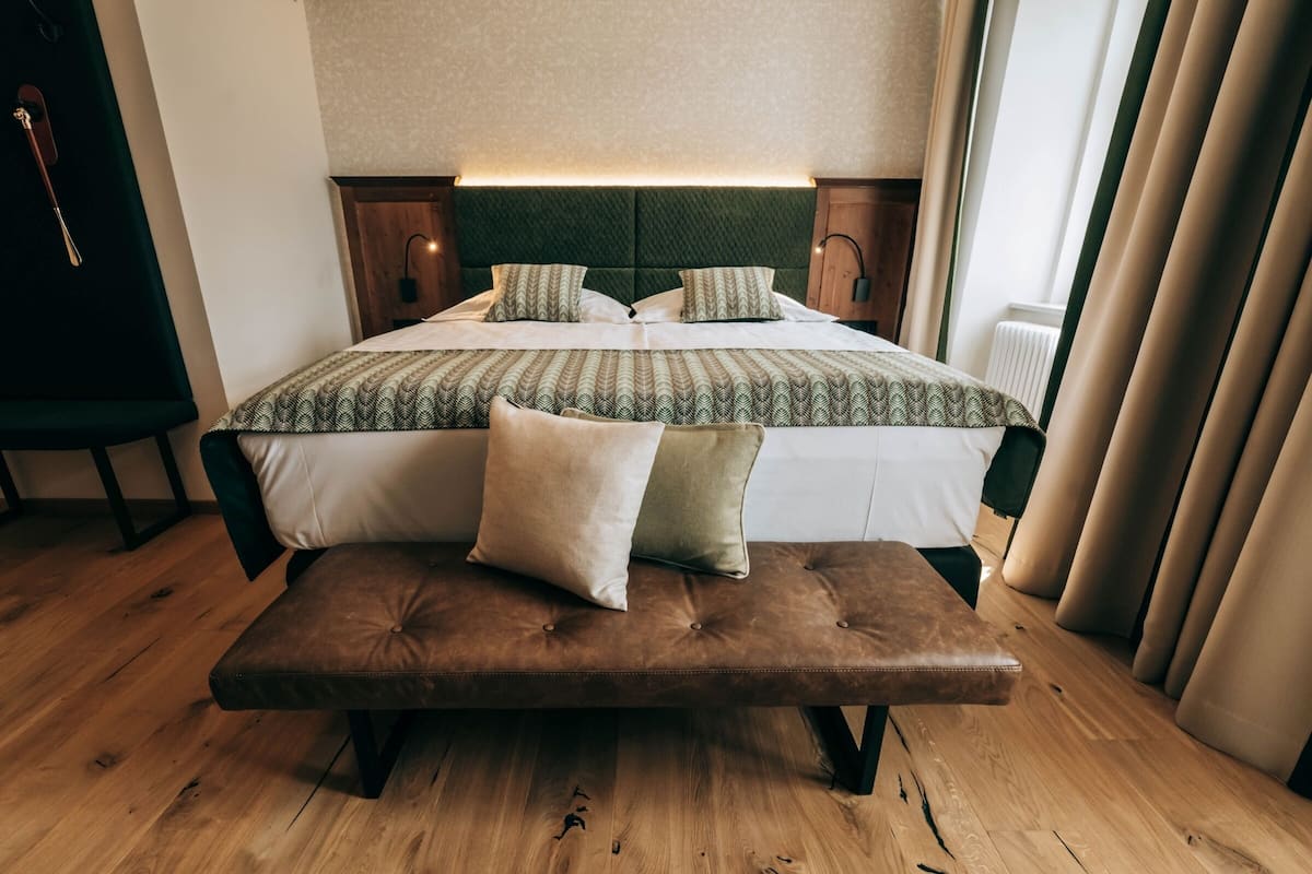 Cama bem arrumada com roupa de cama estampada, um banquinho no rodapé da cama, cabeceira de madeira e uma janela com cortinas.