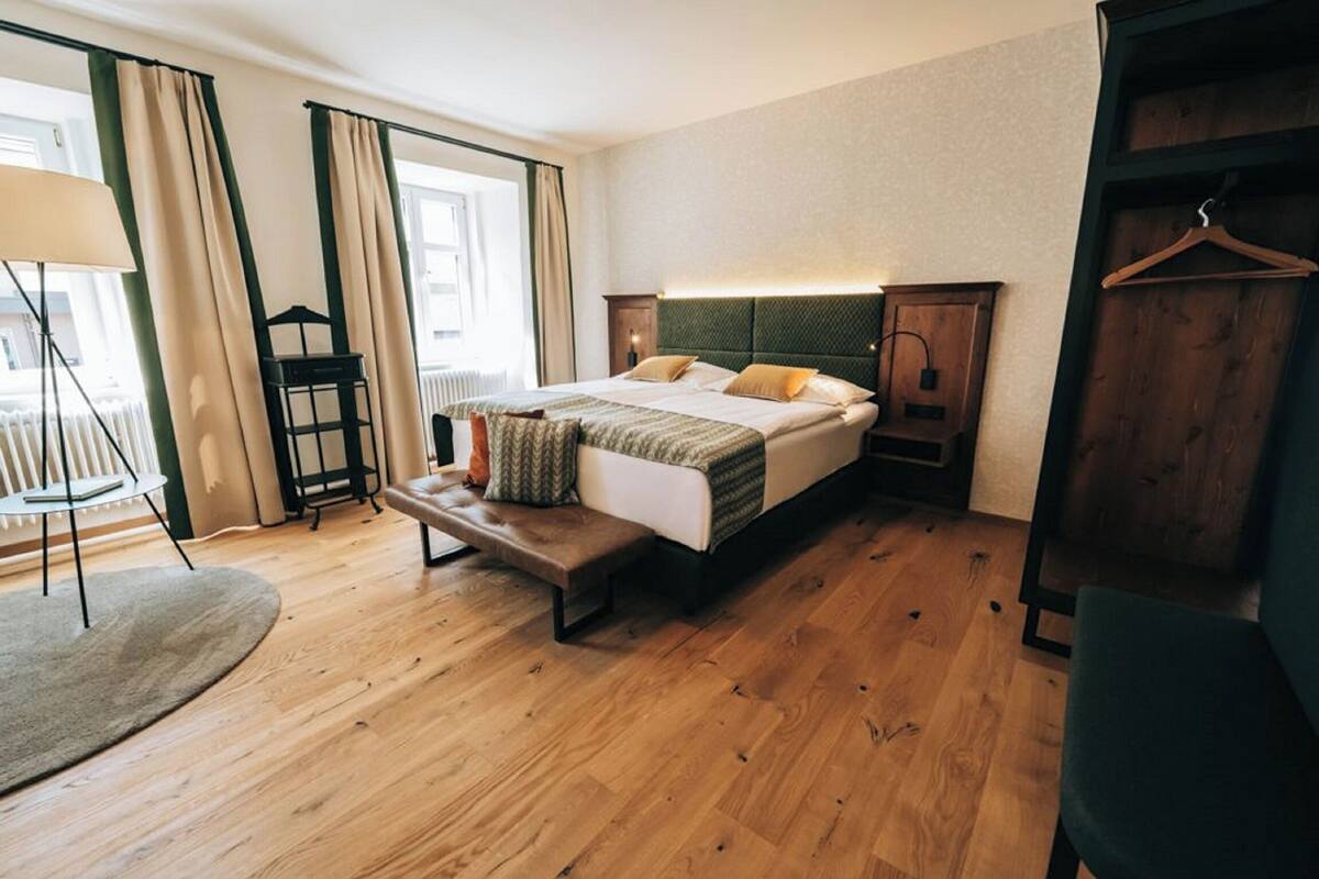 Quarto de hotel com cama, mesas de cabeceira, um banco, guarda-roupa e janela com cortinas.