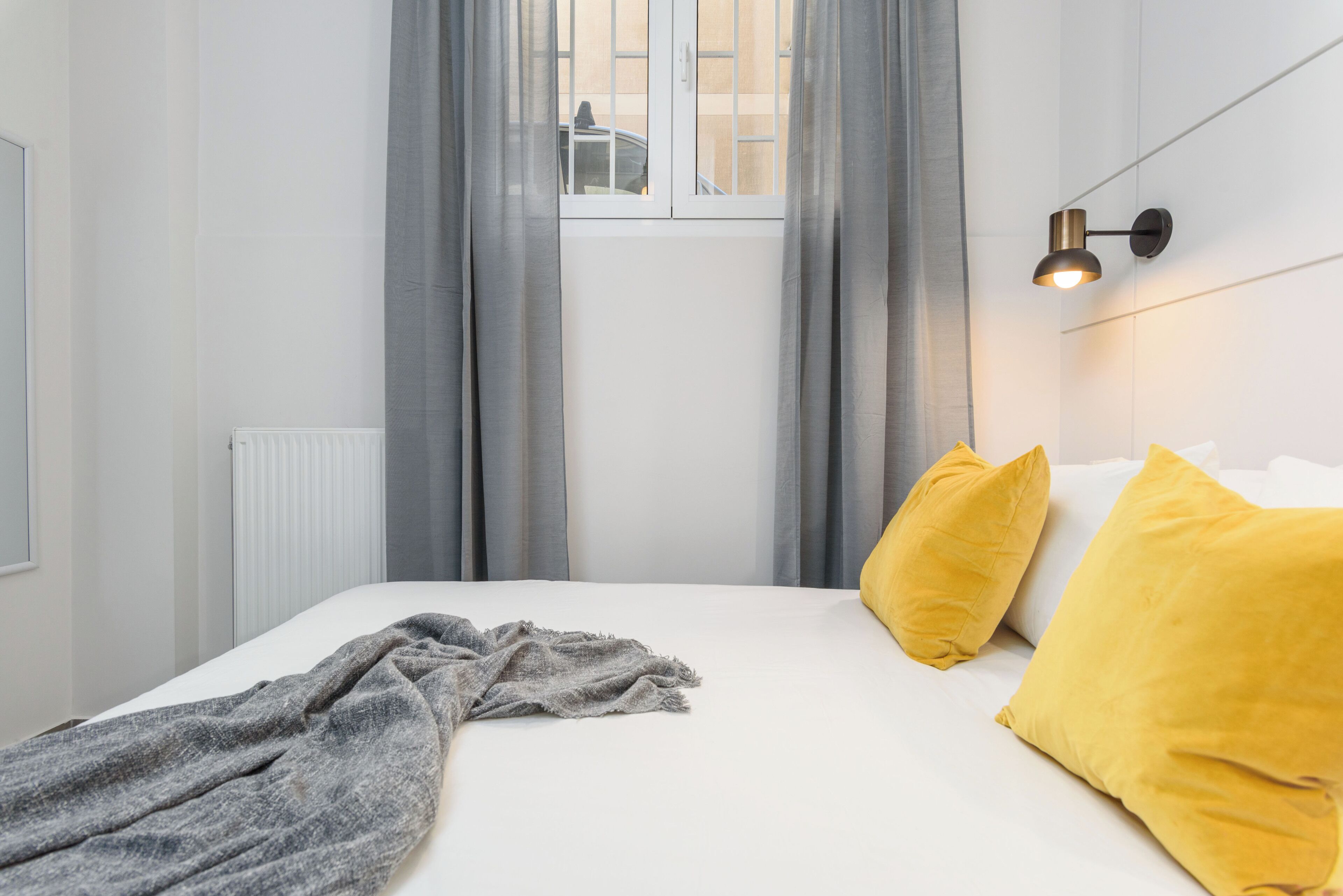 Quarto com cama, cortinas cinzas, janela e luminária fixada na parede.