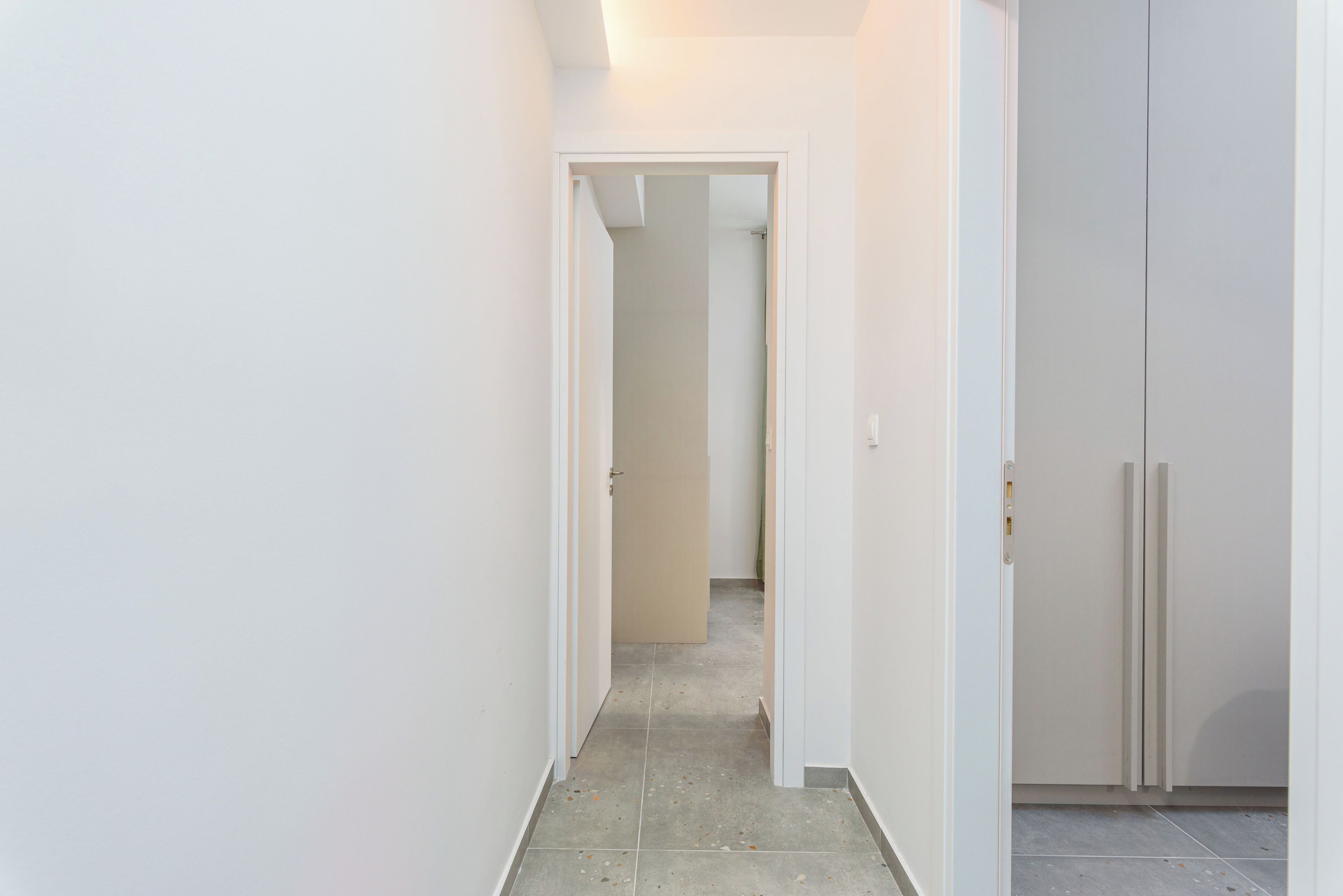 Um corredor estreito com uma porta que dá para um quarto. O corredor possui piso de azulejos e paredes brancas.