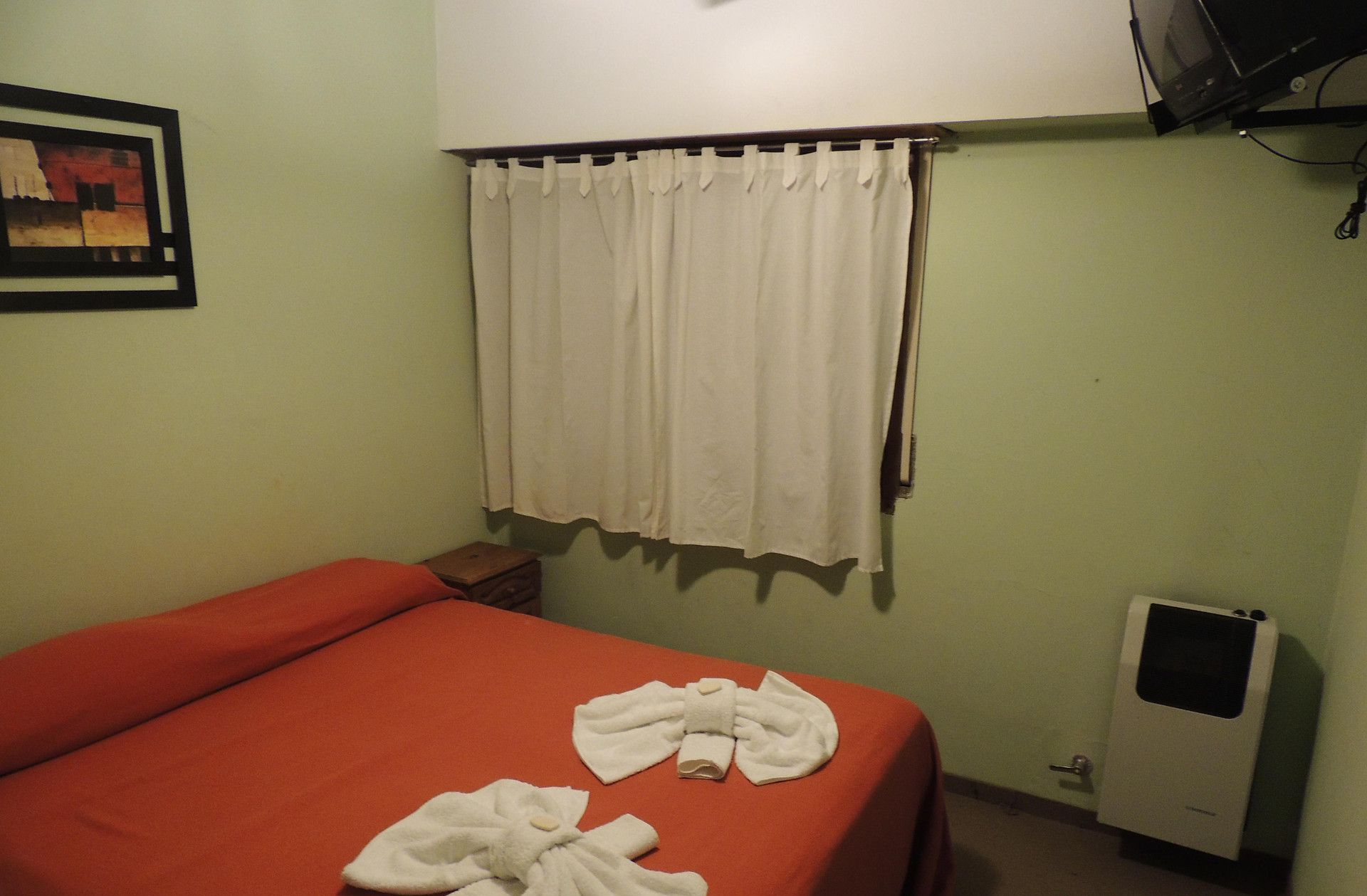Uma cama de solteiro com colcha vermelha e duas toalhas dobradas. Há uma cortina branca em uma janela, uma pequena mesa de cabeceira de madeira e uma televisão sobre um móvel à direita.