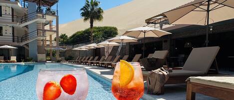 Área à beira da piscina com duas bebidas sobre uma mesa de madeira, cadeiras de descanso e um edifício com piscina ao fundo.