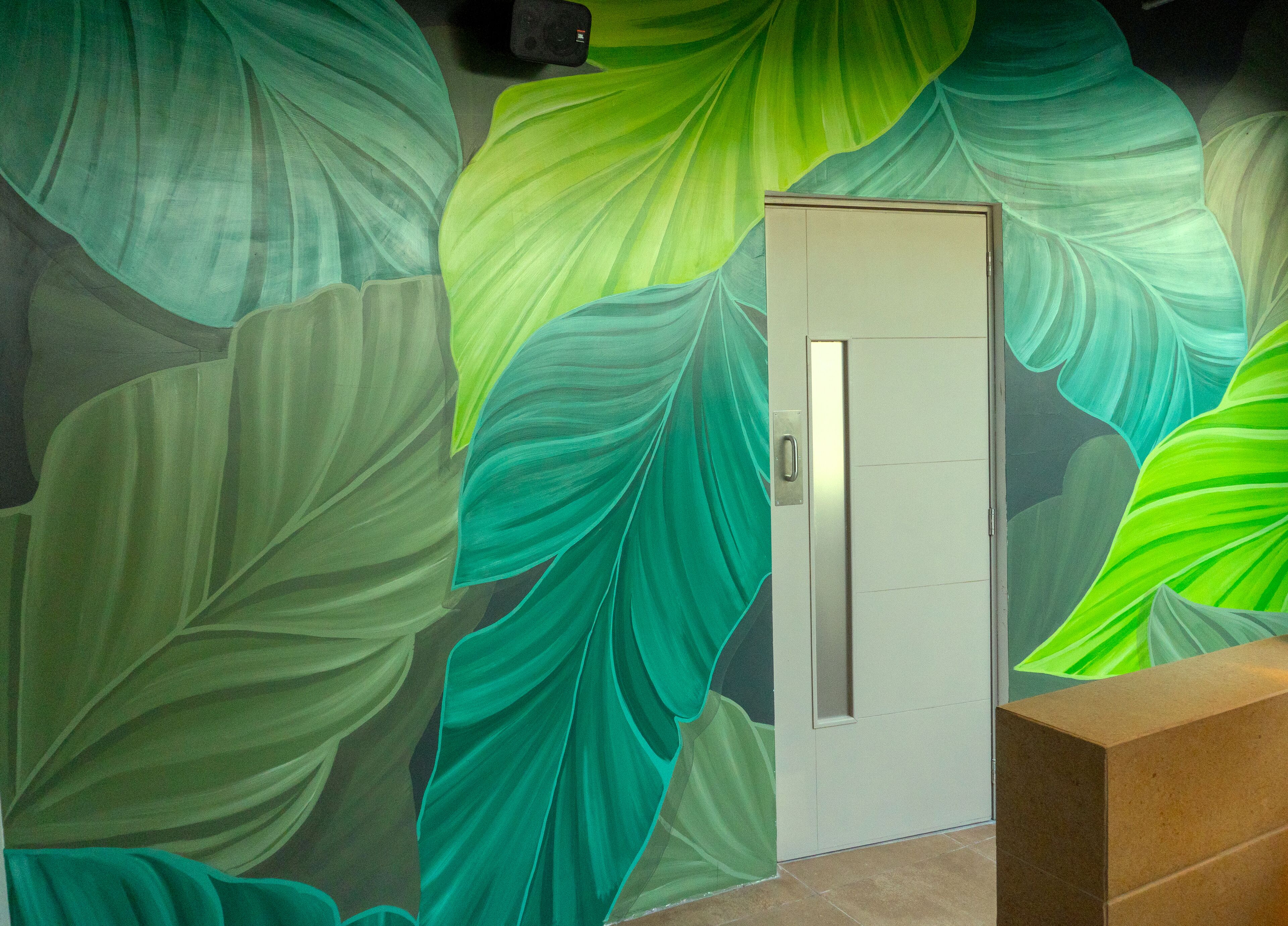 Uma porta com puxador metálico e painel de vidro, em parede com padrões de folhas grandes e abstratas.