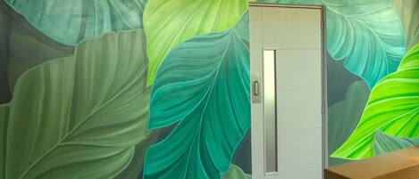 Uma porta com puxador metálico e painel de vidro, em parede com padrões de folhas grandes e abstratas.