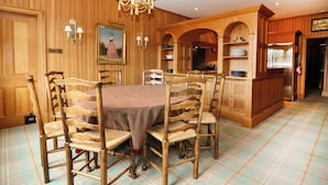 Uma sala de jantar com mesa e cadeiras de madeira, um candelabro e uma área de cozinha com armários de madeira e uma geladeira.