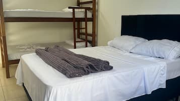 Uma cama de solteiro com colchão branco e cabeceira escura, uma cama beliche com escada de madeira, e uma parede branca com uma pequena janela.