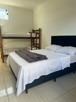 Uma cama de solteiro com colchão branco e cabeceira escura, uma cama beliche com escada de madeira, e uma parede branca com uma pequena janela.