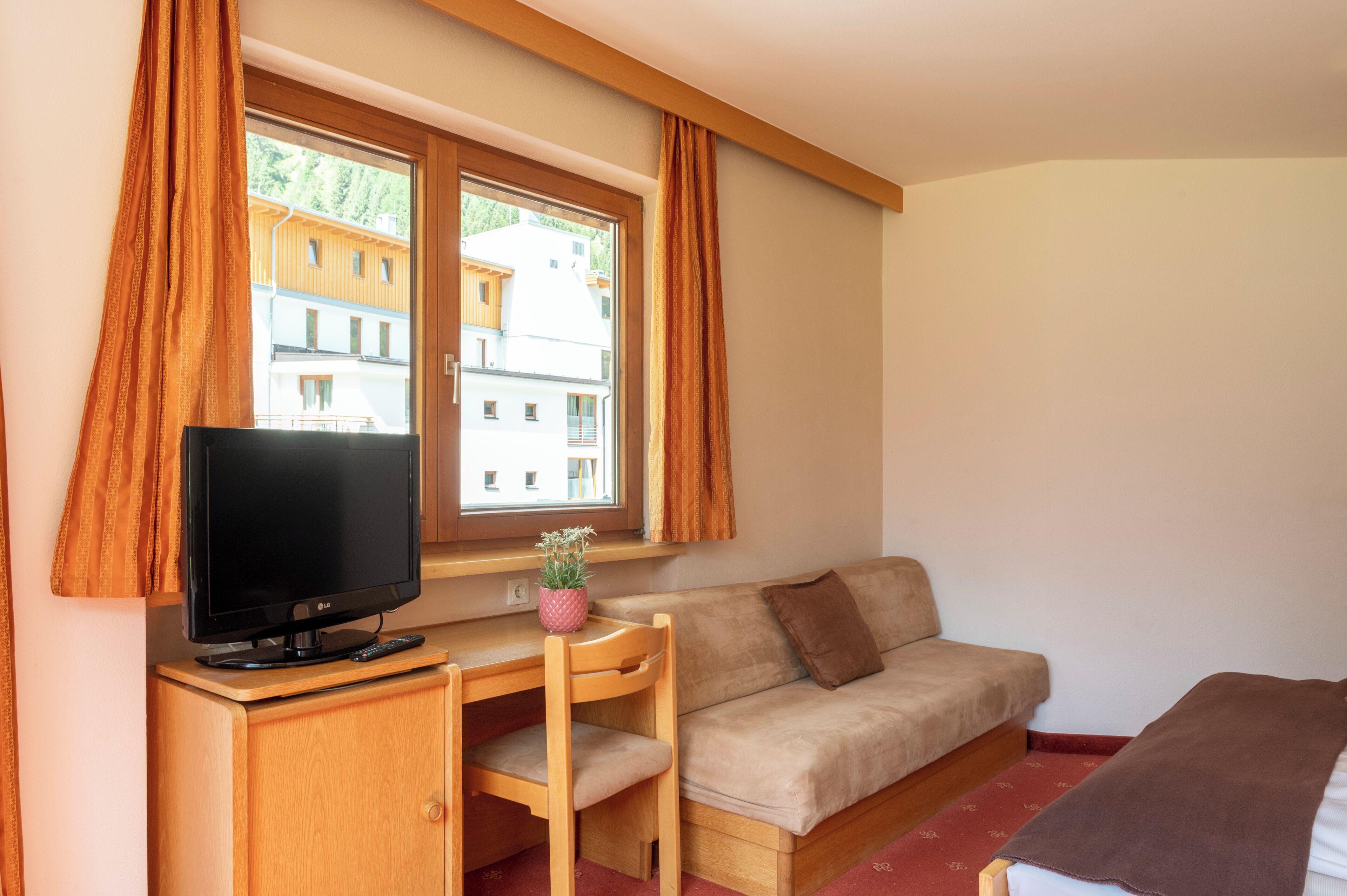 Quarto com televisão sobre uma escrivaninha de madeira, um sofá e uma janela com cortinas.