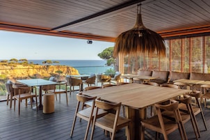 Um deck de madeira com mesas e cadeiras, com vista para a paisagem costeira com falésias e o mar.