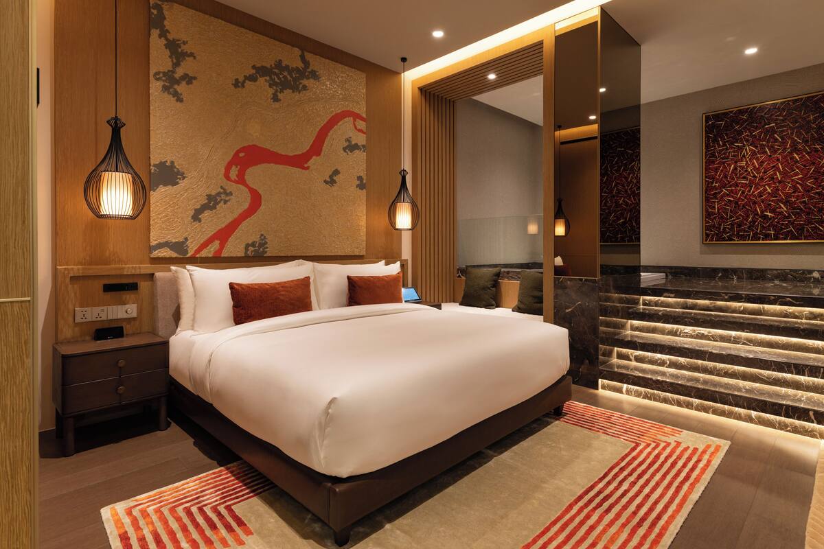 Quarto de hotel moderno com uma cama grande, uma mesa de cabeceira, uma luminária na parede e uma escada com degraus iluminados.