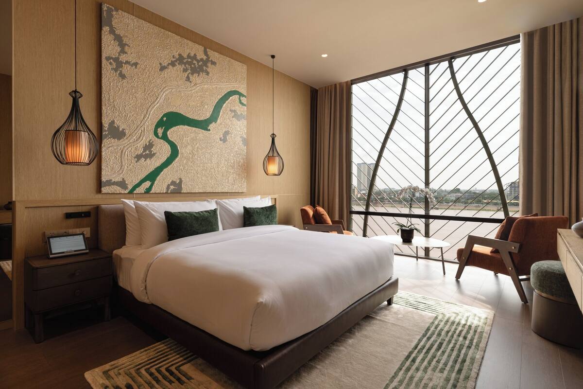 Quarto de hotel moderno com uma cama grande, duas mesas de cabeceira, uma escrivaninha, uma cadeira, vista pela janela ampla e obras de arte na parede.