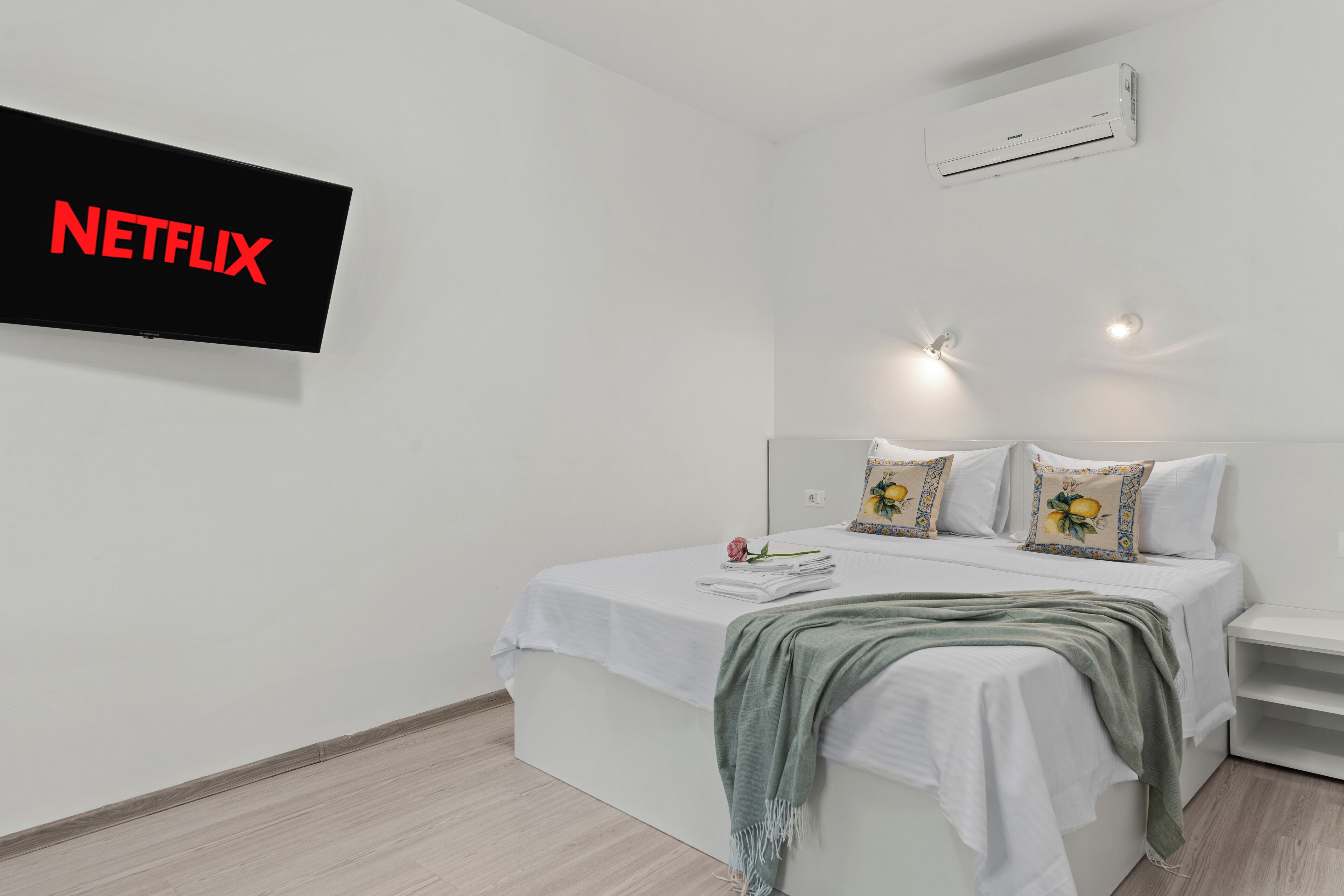Quarto de hotel moderno com televisão de tela plana exibindo o logotipo da Netflix, cama bem arrumada com manta verde e ar-condicionado fixado na parede.