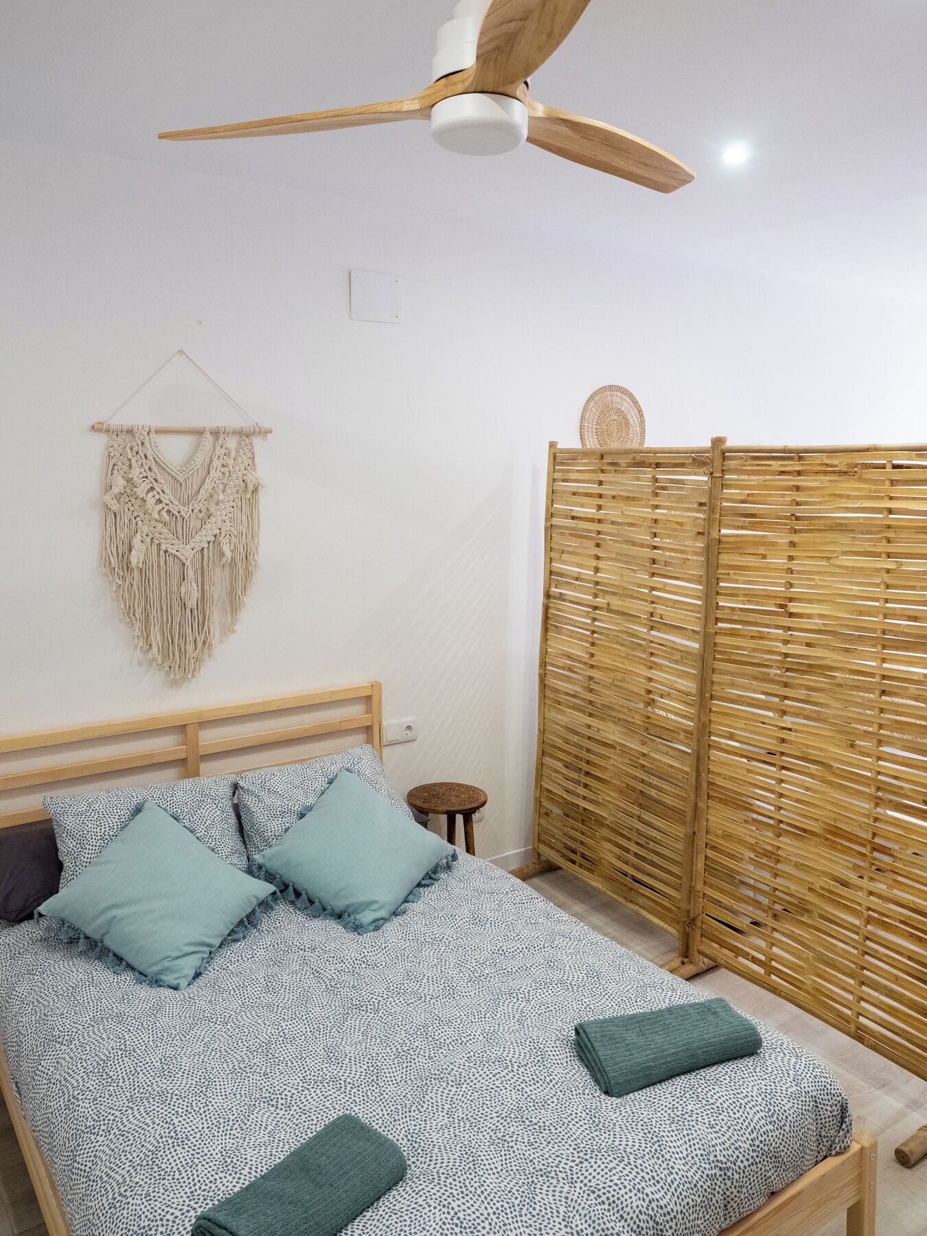 Quarto com cama, ventilador de teto, um pendente de macramé, um divisório de tecido e um banquinho.
