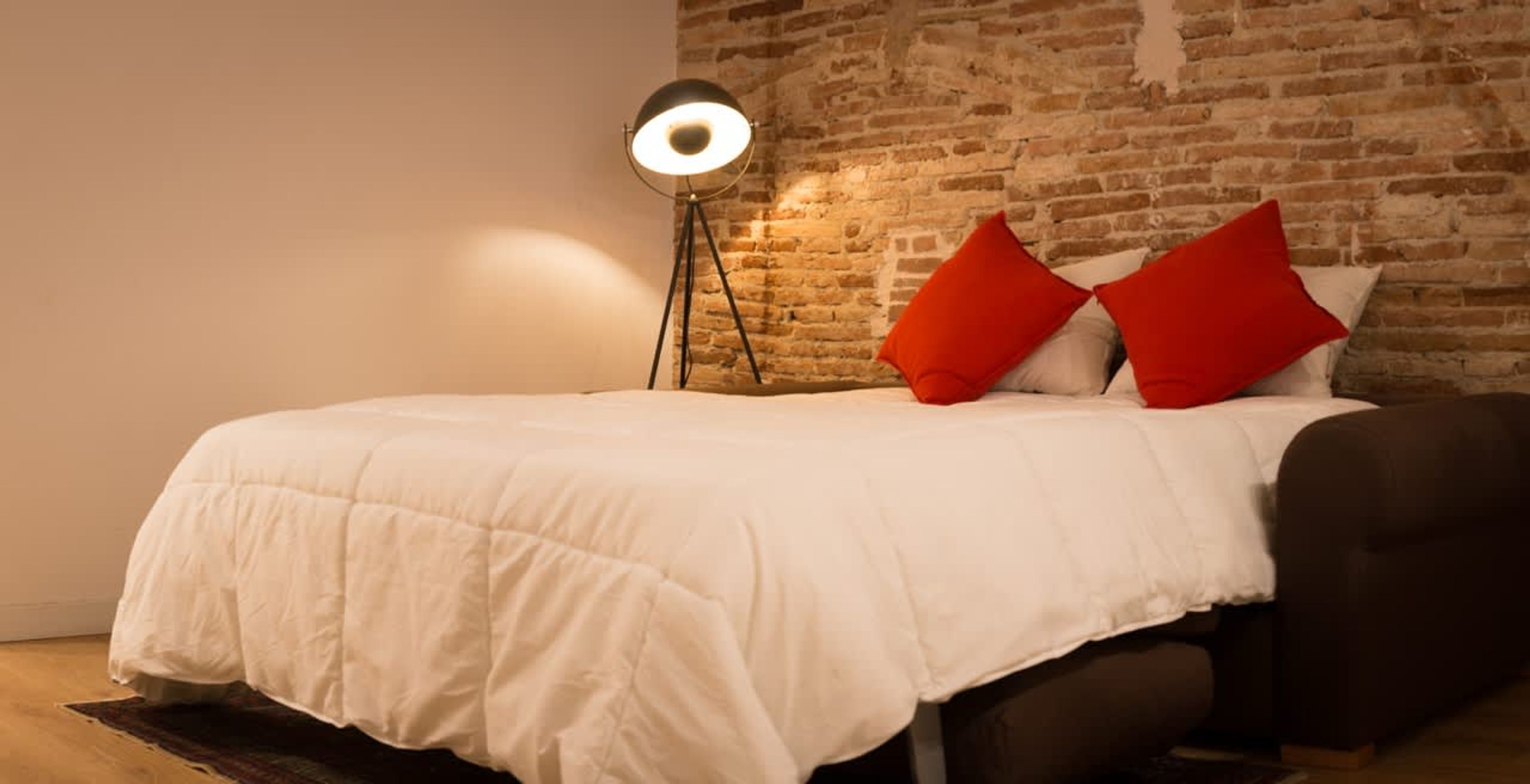 Uma cama com roupa de cama branca, almofadas vermelhas e um abajur com cúpula branca.