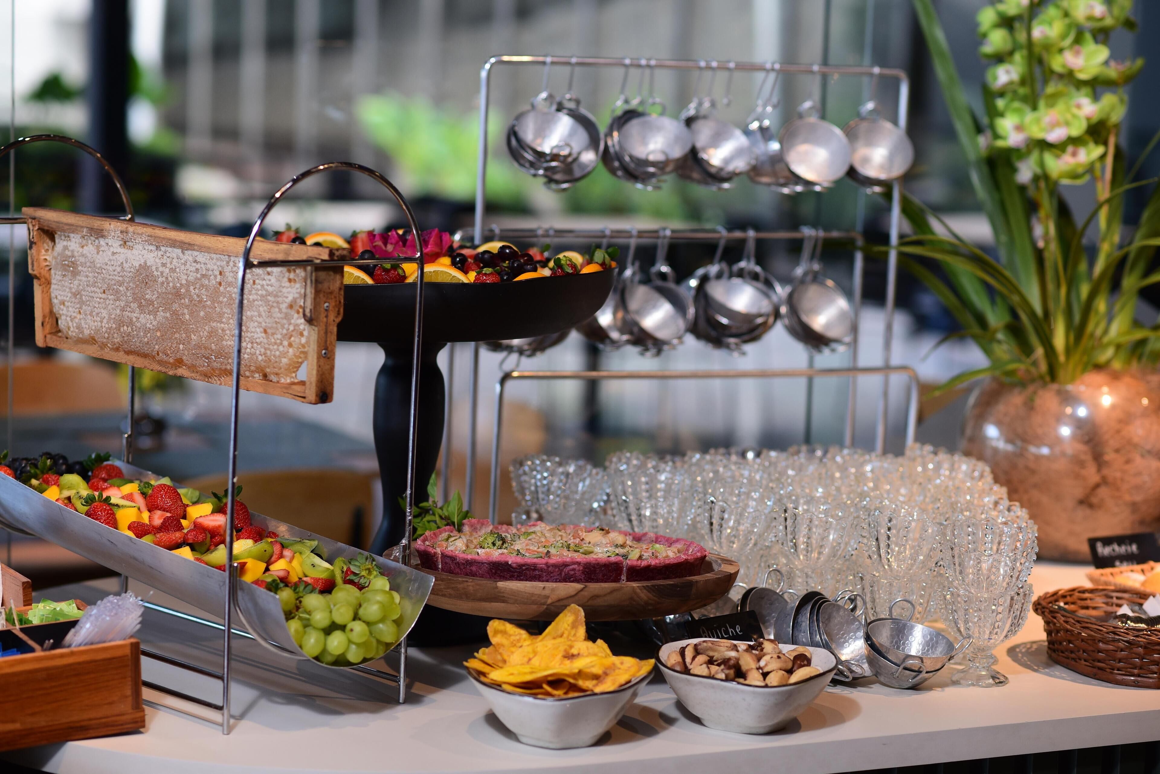 Mesa de buffet com diversos alimentos, uma exposição de frutas e um conjunto de copos.