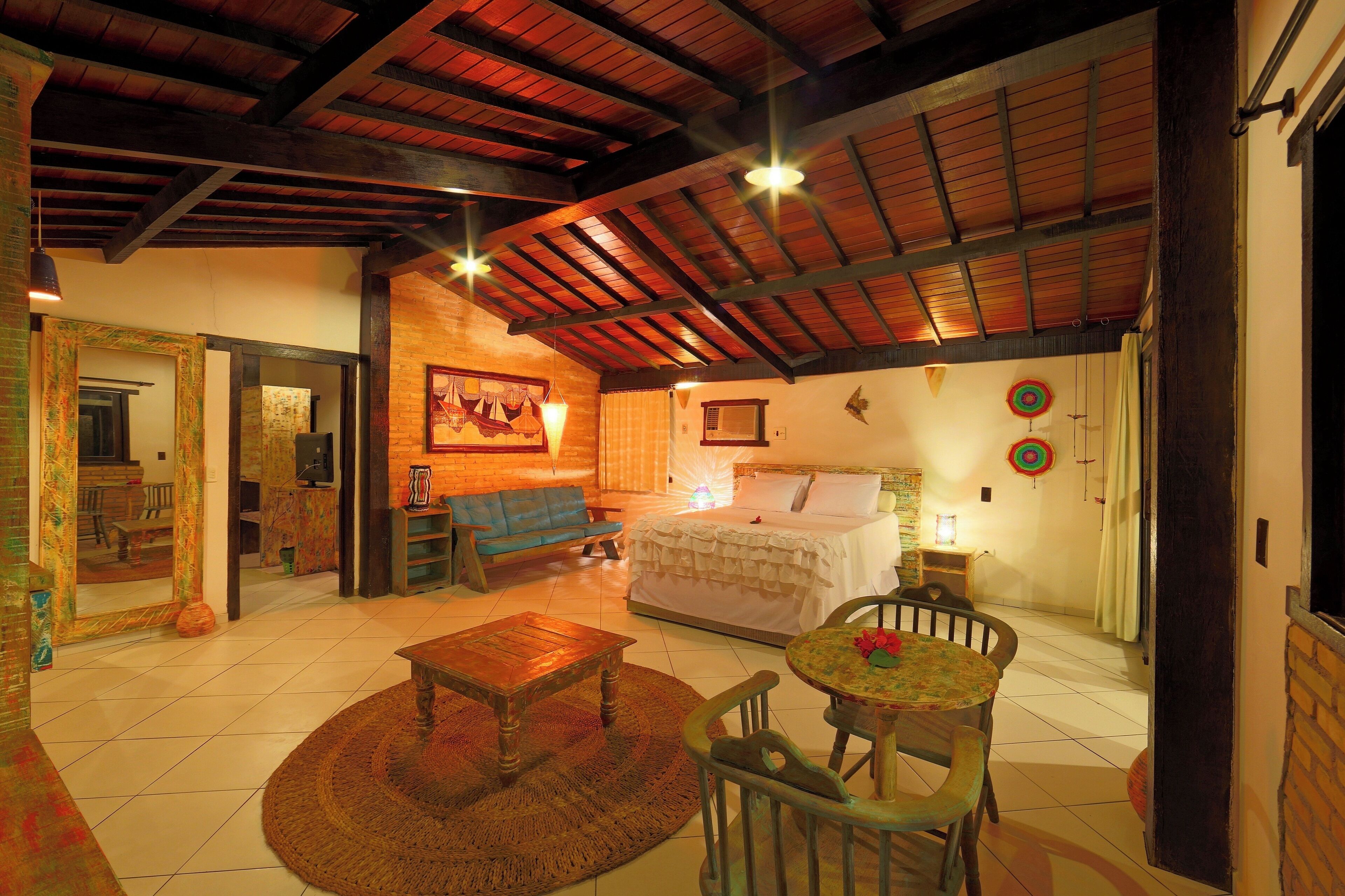 Um quarto aconchegante com cama, sofá, mesa de jantar e mesa de centro. Há um espelho na parede e uma janela com cortinas.