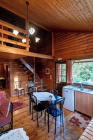 Interior de uma cabana de madeira com área de jantar, cozinha e escada que leva a um andar superior.