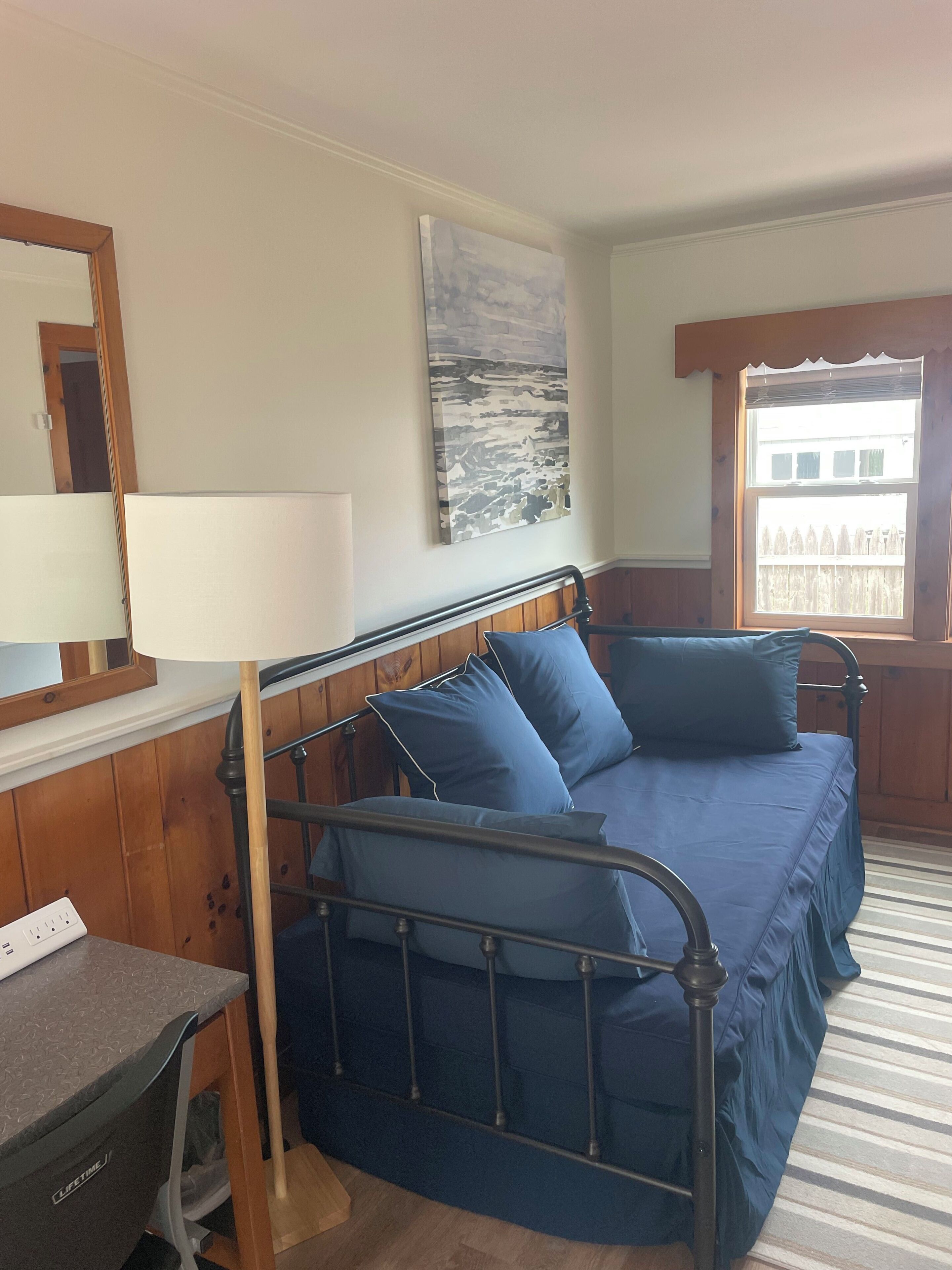 Quarto com cama de estrutura metálica, mesa de cabeceira com luminária, janela com vista para uma cerca e um quadro pendurado na parede.