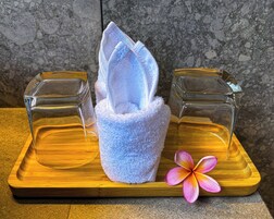 Uma bandeja de madeira com duas garrafas de vidro transparente e uma toalha branca com uma flor rosa.