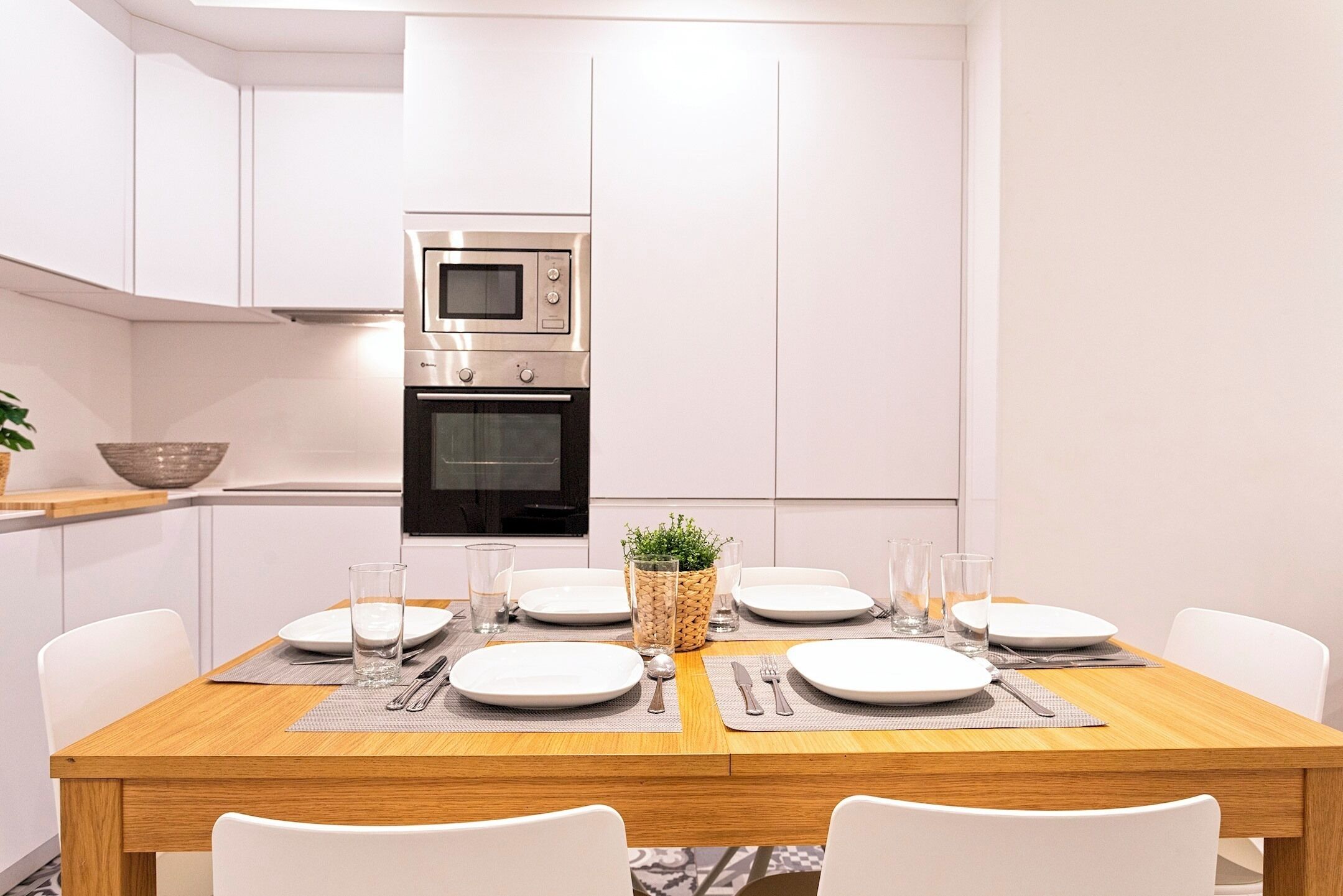 Uma cozinha moderna com armários brancos, forno embutido e mesa de jantar para quatro pessoas, com pratos, talheres e copos brancos.