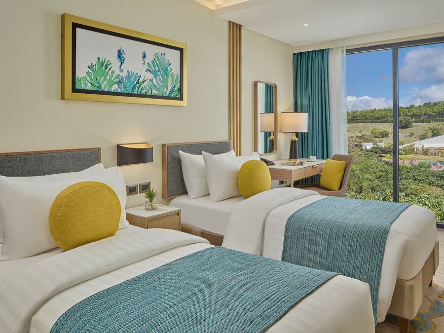 Quarto de hotel com duas camas, uma secretária, uma cadeira, uma janela grande com vista para áreas verdes e uma obra de arte de cavalo-marinho emoldurada na parede.