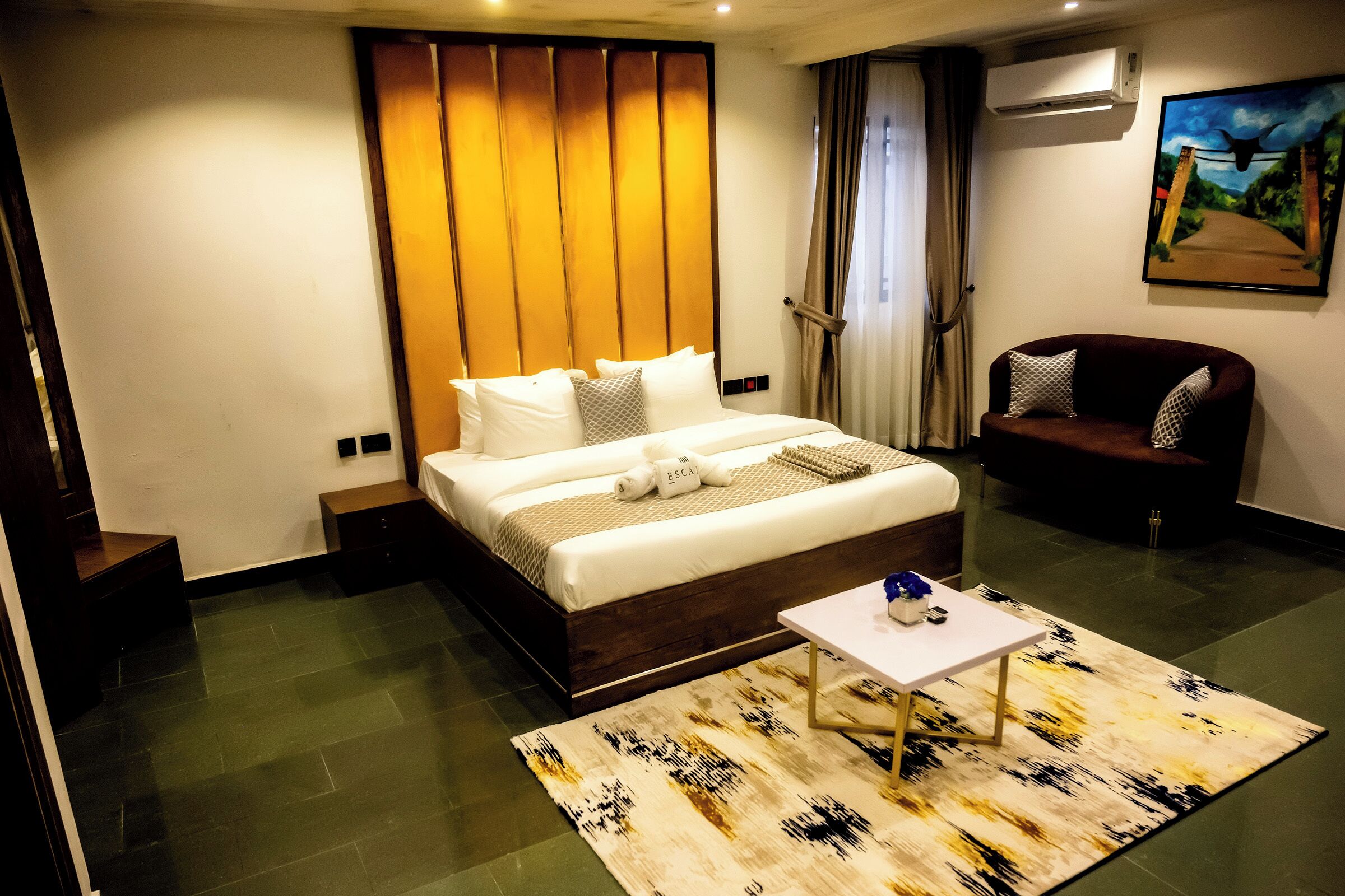 Quarto de hotel moderno com uma cama grande, um sofá pequeno, uma mesa de centro e um quadro na parede.