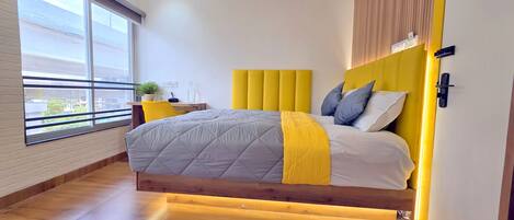 Quarto moderno com uma cama grande, cabeceira amarela e vista para o exterior por uma janela ampla.