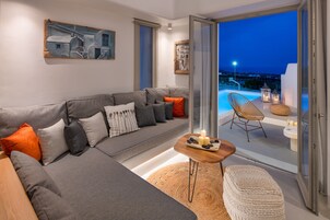 Uma sala de estar moderna com um sofá cinzento, uma pequena mesa de madeira com velas e vista para uma piscina e céu noturno.